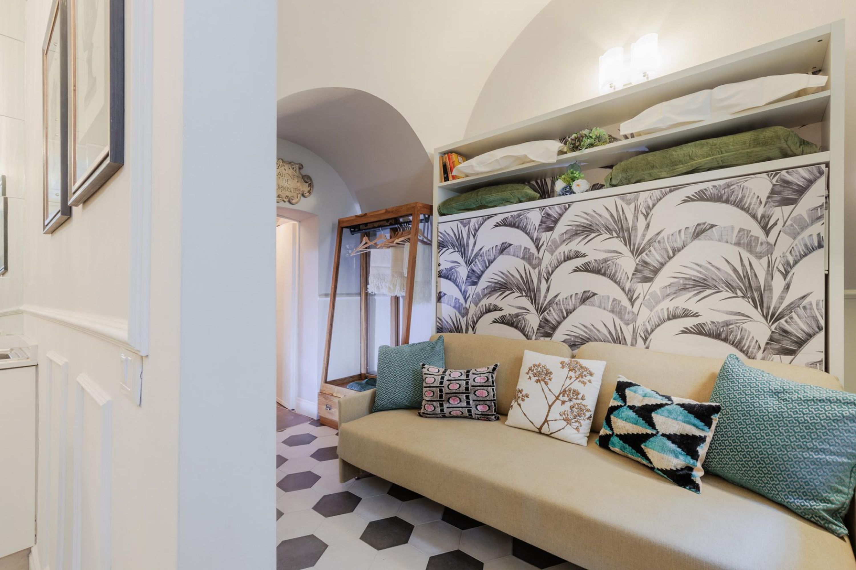 Uma sala de estar acolhedora com um sofá, almofadas estampadas e uma parede com design de folhagem tropical.