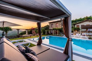 Área de piscina com espreguiçadeiras, um espaço coberto para relaxar e vista para um edifício de pedra e vegetação exuberante.