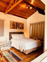 Quarto com teto de madeira, cama com roupa de cama branca, uma cadeira branca, um tapete listrado e duas pinturas penduradas na parede.
