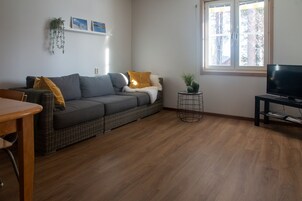 Uma sala de estar com um sofá cinzento, uma mesa redonda pequena, uma televisão sobre um móvel e uma janela com vista para árvores.