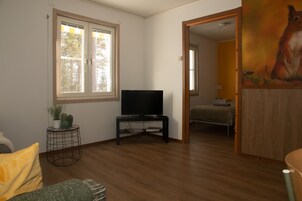 Sala de estar com televisão em um móvel, uma pequena mesa lateral com uma planta, uma janela com vista para árvores e um quadro na parede.