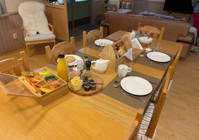 Mesa de jantar posta com pratos, talheres, temperos e uma cesta de pão, num ambiente acolhedor com móveis de madeira e uma área de estar confortável.