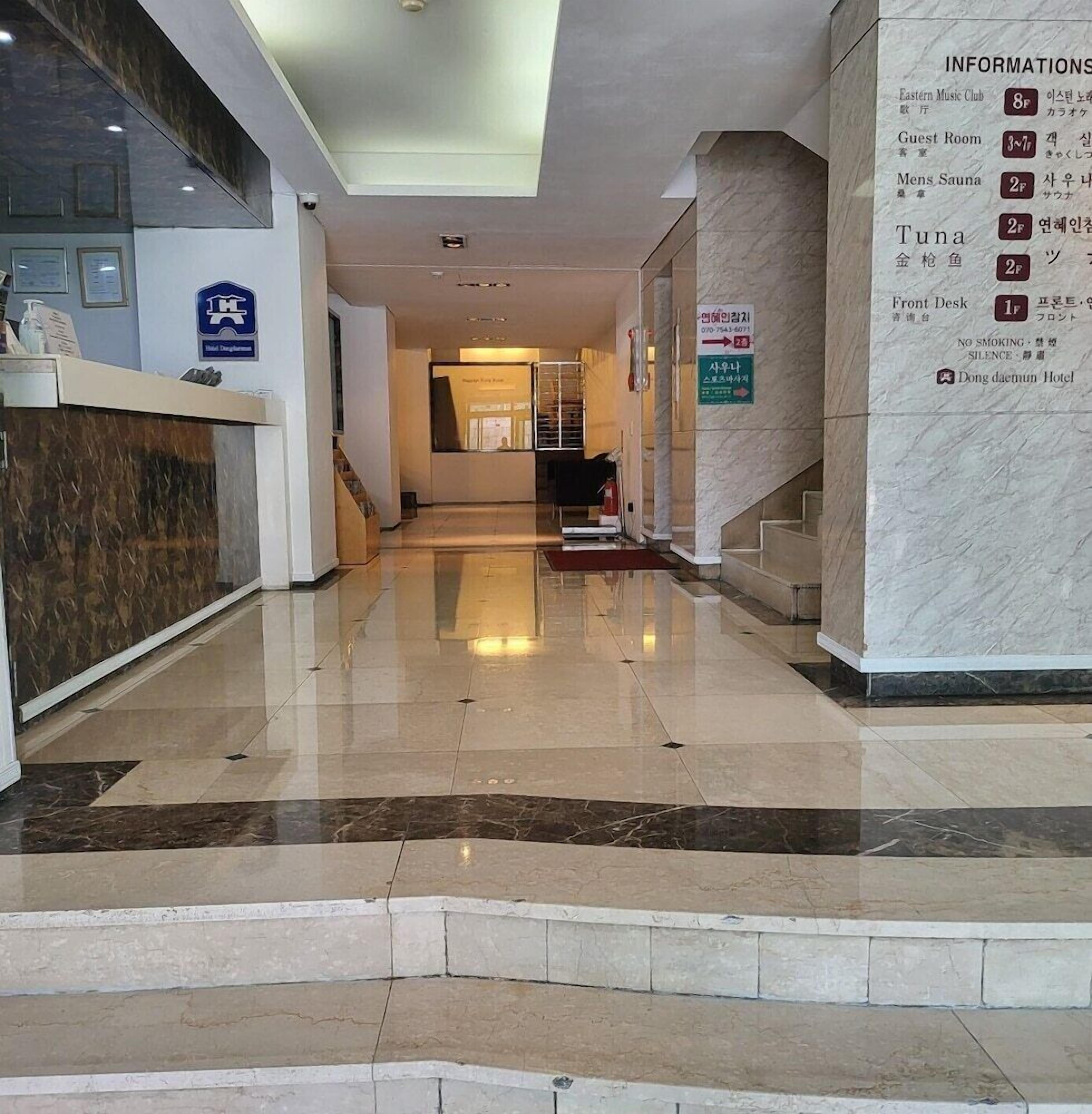 Uma área de recepção de hotel com piso de mármore, balcão de atendimento e escada que leva aos andares superiores.