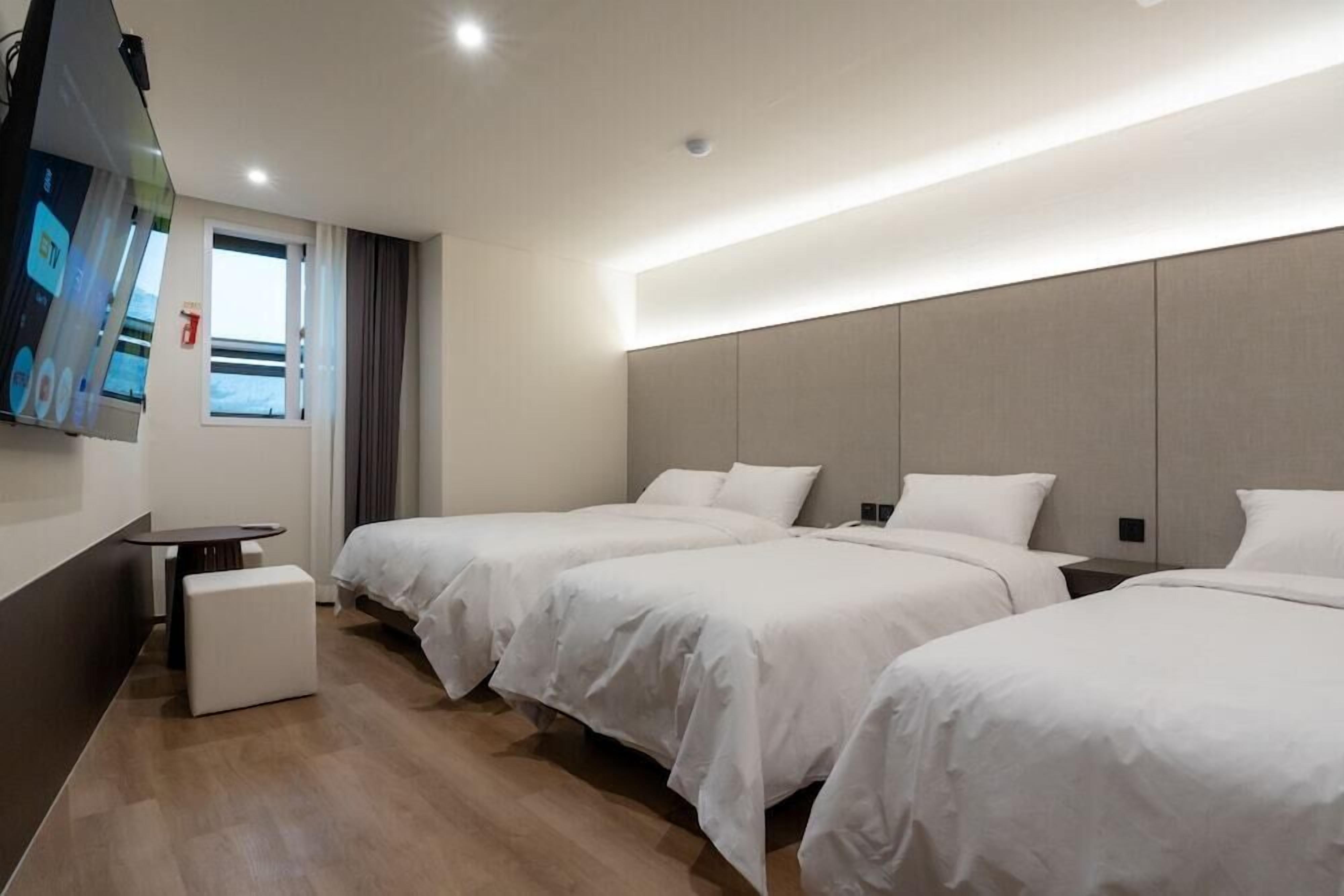 Quarto de hotel com três camas, uma mesa pequena, uma televisão e uma janela com cortinas.