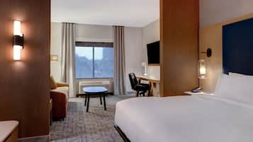 Quarto de hotel com uma cama grande, uma escrivaninha com cadeira, uma mesinha e uma janela com cortinas.