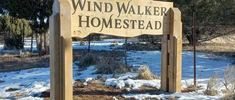 Uma placa de madeira com o texto 'Wind Walker Homestead' em um cenário rural com neve no chão e árvores ao fundo.