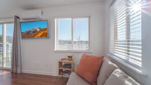 Uma sala de estar com sofá, televisão fixada na parede, uma pequena mesa de apoio de madeira e janela com persianas.