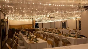 Salão de banquetes com mesas montadas para um evento formal, decorado com luzes de fada e toalhas de mesa brancas.