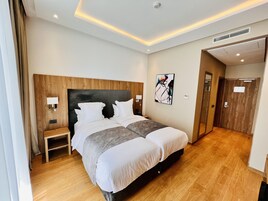 Quarto de hotel moderno com uma cama grande, abajures, uma cabeceira de madeira, um quadro na parede e uma porta que dá para outro quarto.