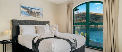 Quarto com cama grande, mesa de cabeceira, pintura na parede e vista para um lago através de uma porta de vidro.