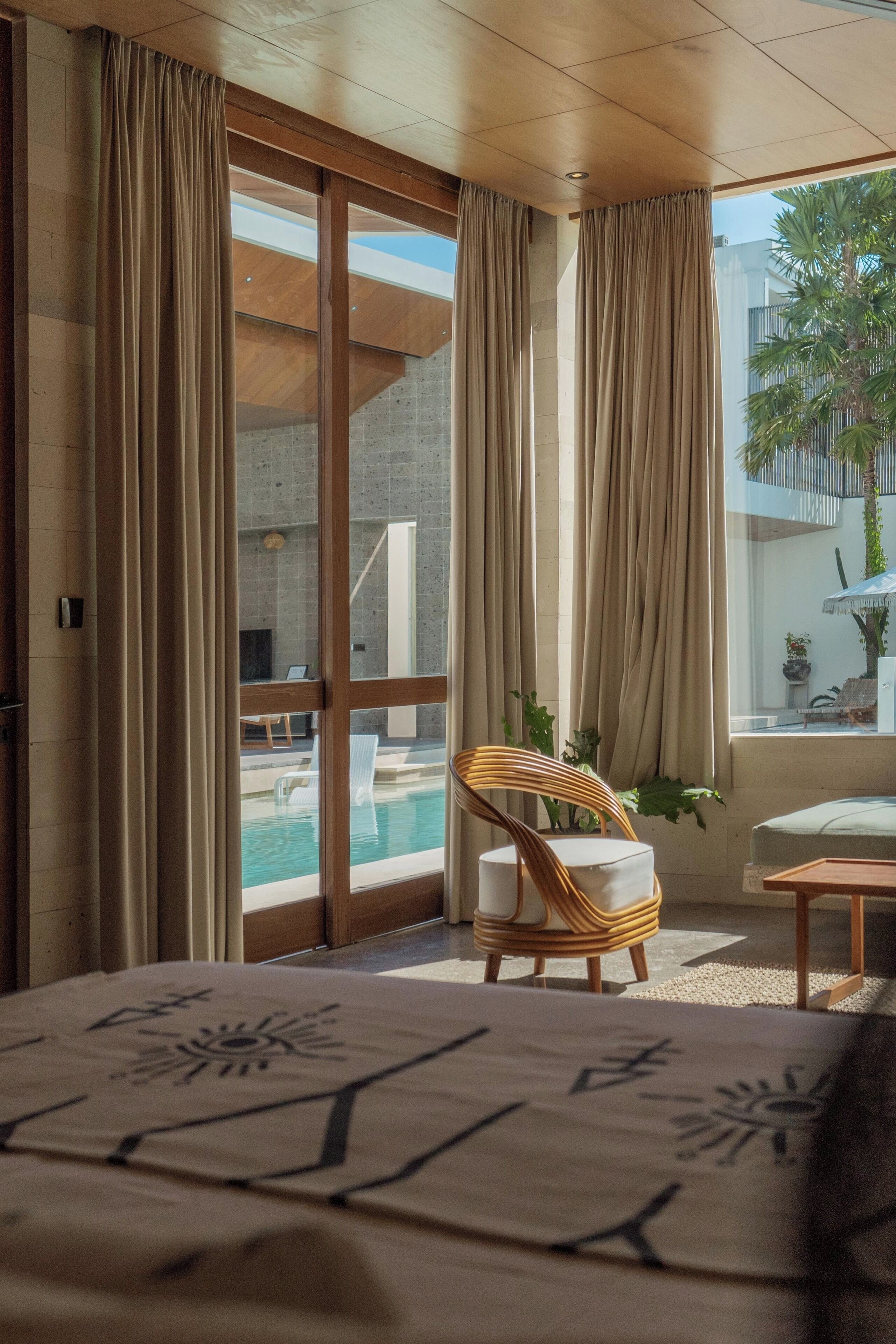 Quarto de hotel moderno com uma grande porta de correr de vidro que dá para uma área de piscina, um espaço de estar confortável com uma poltrona e mesa, e uma cama com colcha estampada.