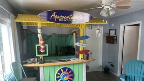 Um espaço de bar pequeno e colorido com tema náutico, exibindo uma placa com a inscrição "Aguaventure Boat Club", com um balcão de madeira, cadeiras e placas indicativas para diversos destinos.