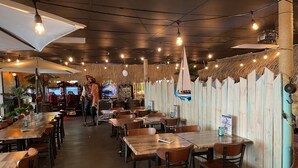 Interior de restaurante com mesas e cadeiras de madeira, área de bar e bar tiki.