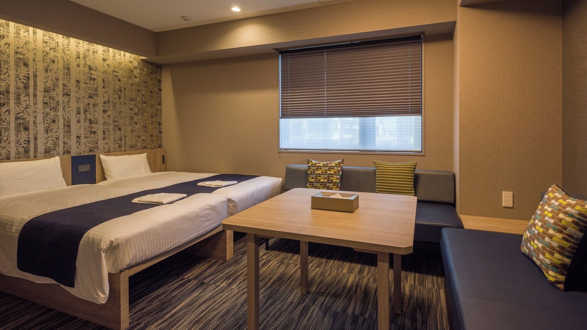 Quarto de hotel com duas camas, uma mesa pequena, um sofá, uma janela com persianas e uma parede estampada.