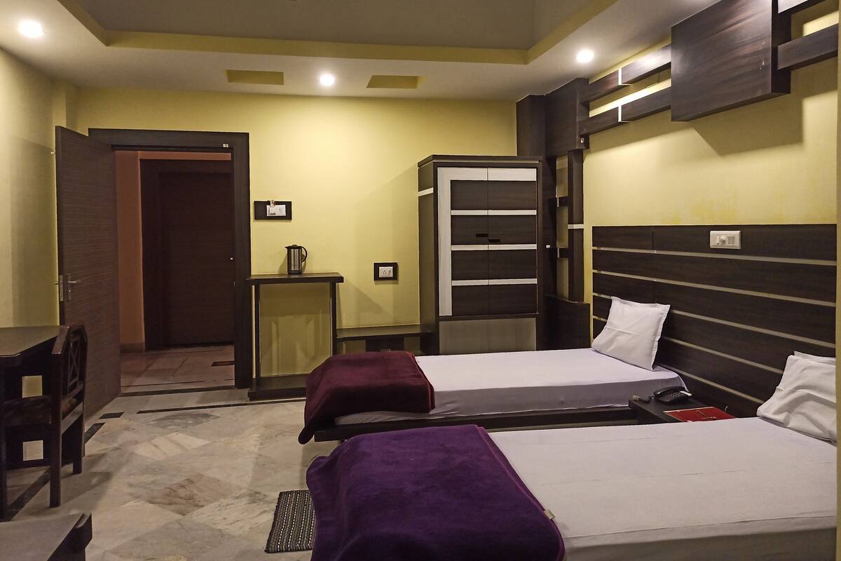 Quarto de hotel com duas camas, uma escrivaninha, uma cadeira, um guarda-roupa e um ventilador de teto.