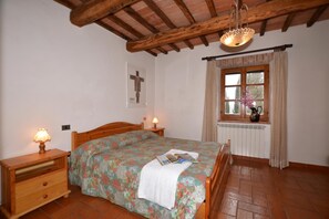Quarto com cama de madeira, mesa de cabeceira com abajur, janela com cortinas e uma cruz na parede.