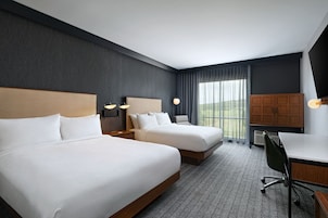 Quarto de hotel com duas camas, uma escrivaninha e uma cadeira. Há duas luminárias na parede e uma janela grande com cortinas.