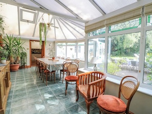 Uma sala de jantar iluminada pelo sol, com uma mesa grande e cadeiras, rodeada por plantas em vasos e amplas janelas com vista para o exterior.