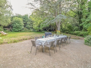 Uma área de refeições ao ar livre com uma mesa posta para seis pessoas, rodeada por cadeiras de vime, em um jardim exuberante.