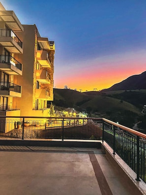 Um edifício com vários andares, varandas, um terraço pavimentado e vista para uma paisagem montanhosa ao entardecer.