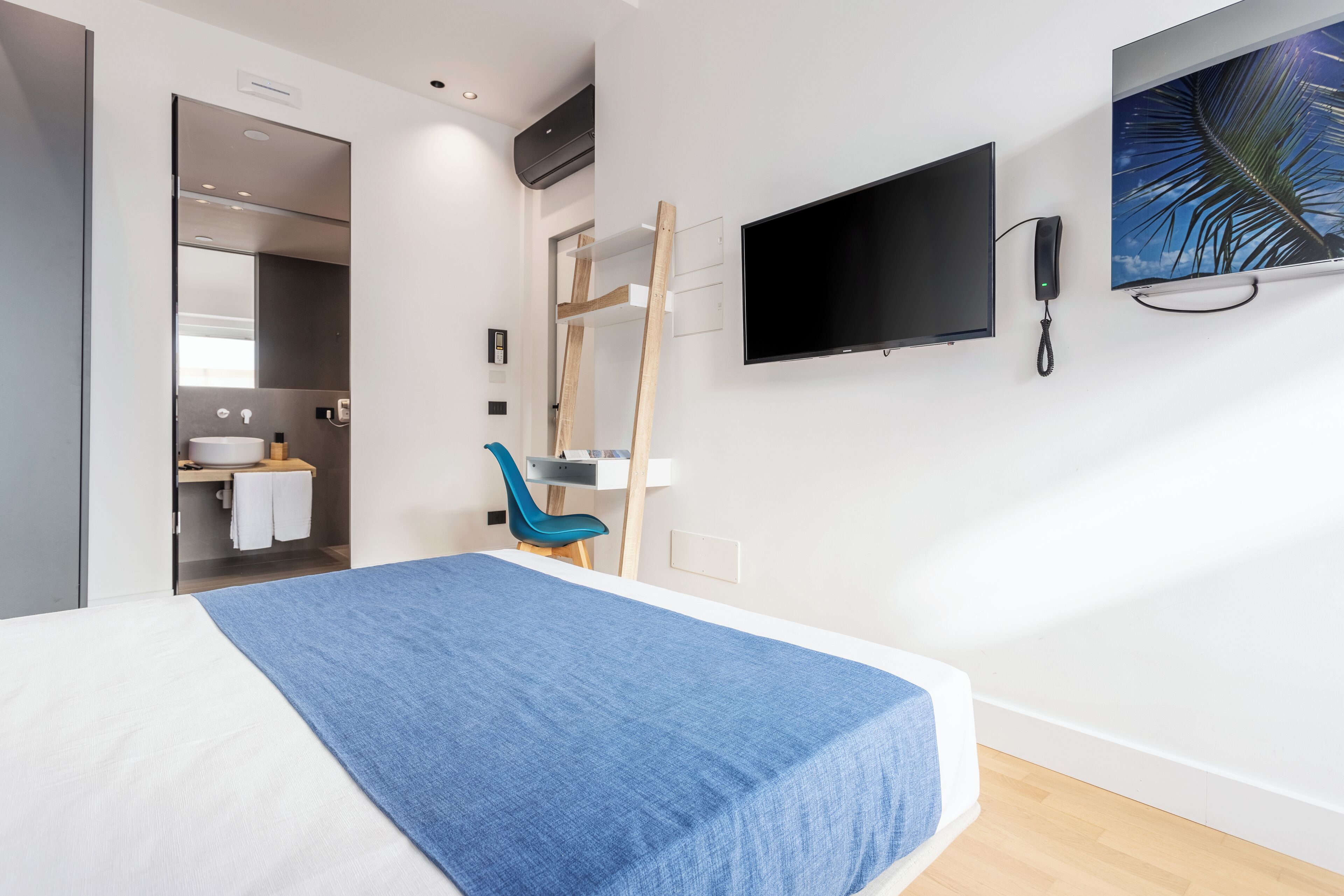 Quarto de hotel moderno com cama, escrivaninha, cadeira, televisão e banheiro visível por uma porta aberta.