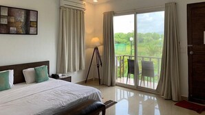 Quarto de hotel com cama, luminária, tripé e vista para o verde pelas janelas.