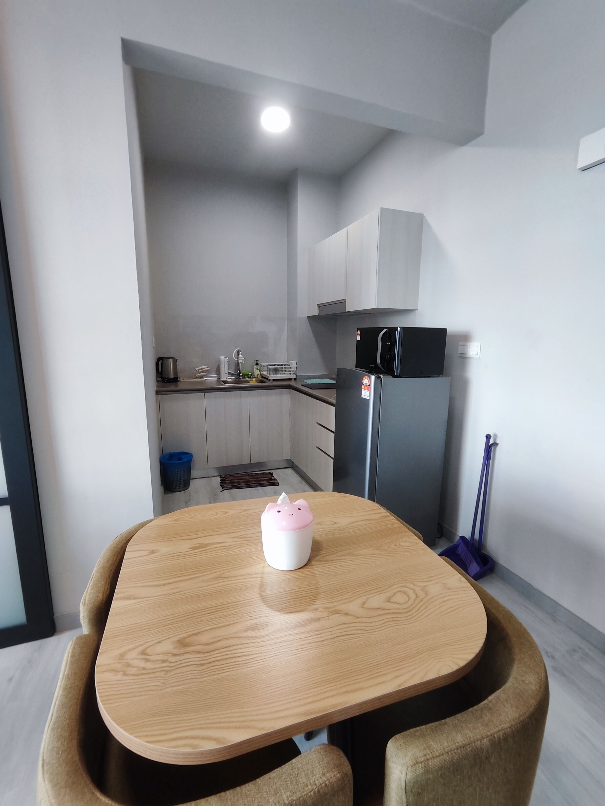 Uma cozinha moderna com micro-ondas, frigorífico e pia. Há uma mesa de jantar de madeira com um recipiente em forma de porco rosa.