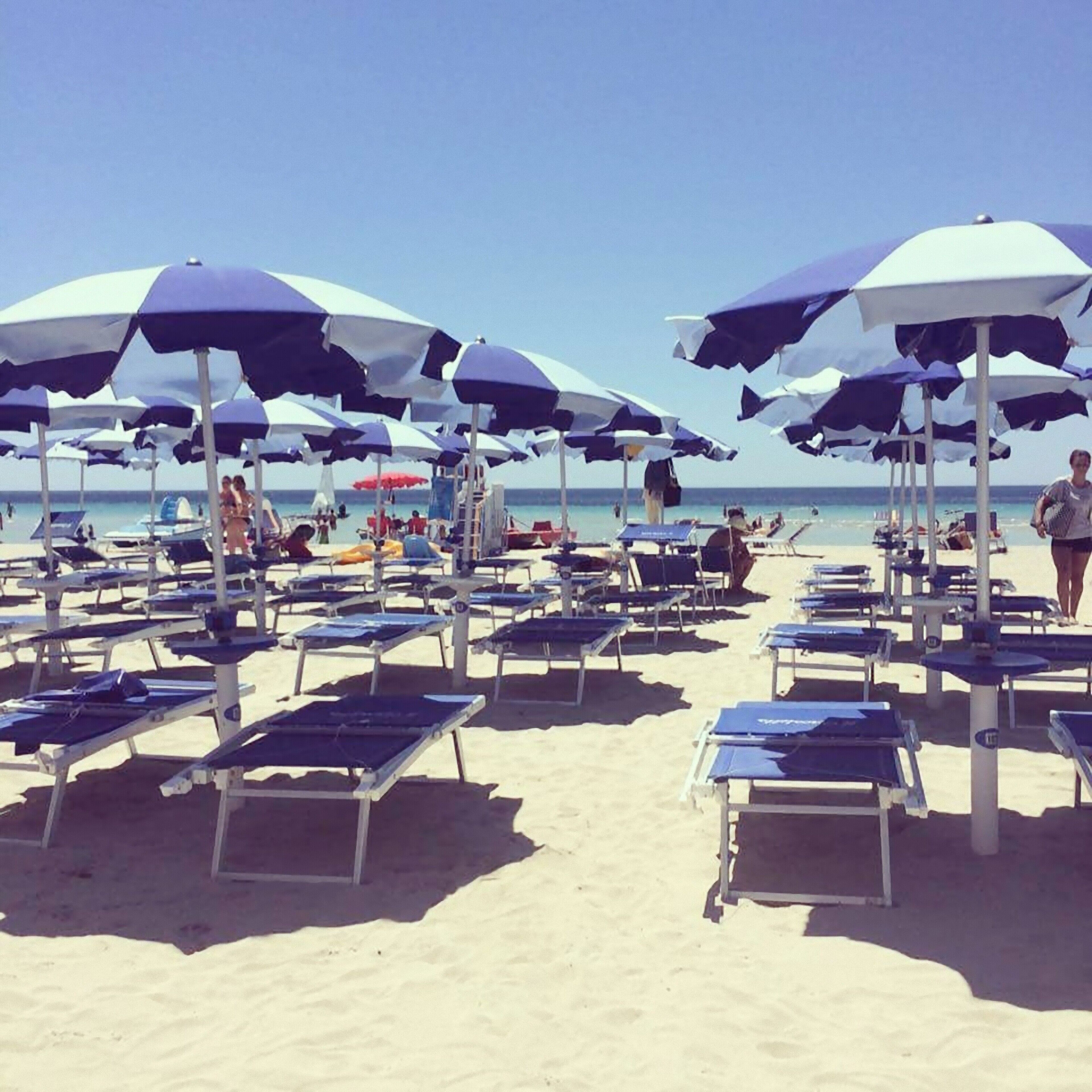 Uma praia com fileiras de espreguiçadeiras e guarda-sóis, céu claro e o oceano ao fundo.
