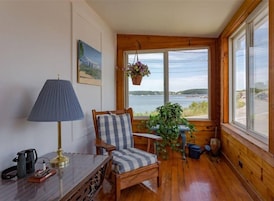 Um quarto de cabana acolhedor com paredes de madeira, vista para um lago, uma poltrona com almofadas listradas, uma mesa com abajur e uma planta pendente.