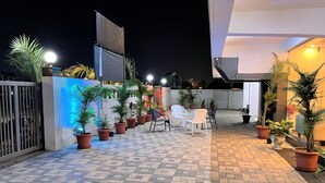 Uma área de pátio externa bem iluminada, com piso de azulejos, plantas em vasos e mobiliário de exterior.