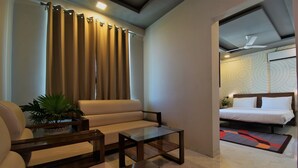 Quarto de hotel moderno com sofá, mesa de centro de vidro, mesa lateral de madeira e cama com edredom branco.