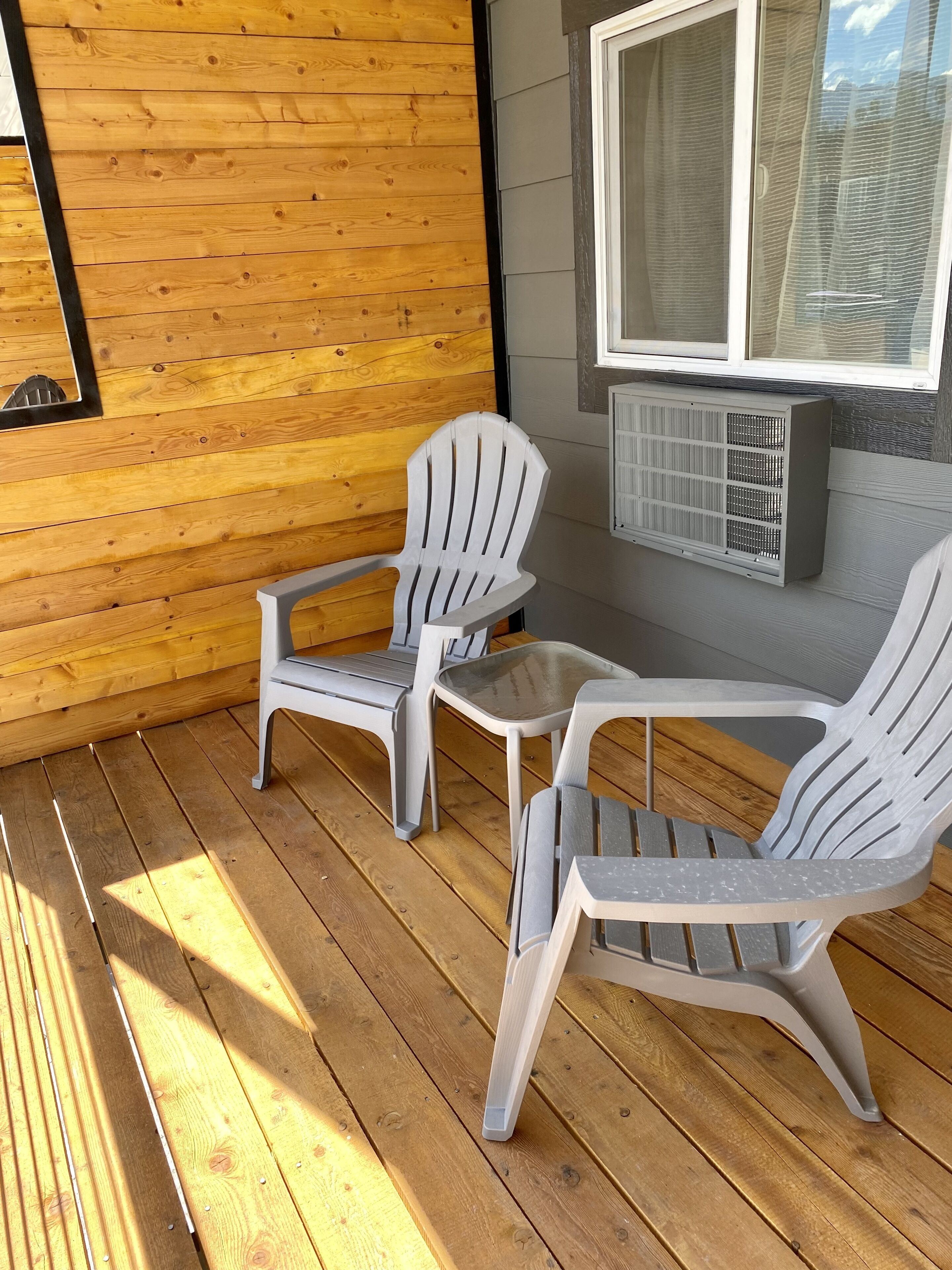 Um deck de madeira com duas cadeiras Adirondack e uma mesa pequena, uma janela com vista e um ar condicionado fixado na parede.