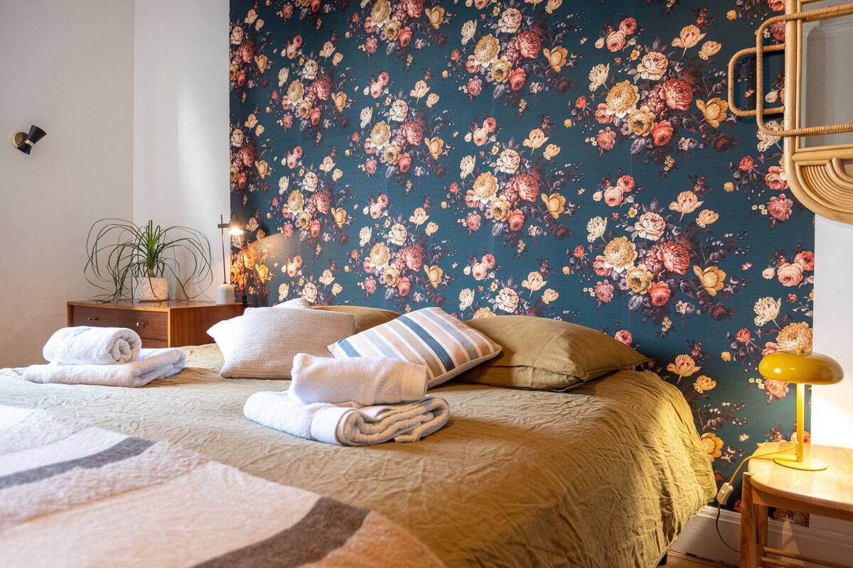 Quarto com papel de parede floral, cama com várias almofadas, mesa de cabeceira com abajur e um vaso de planta.