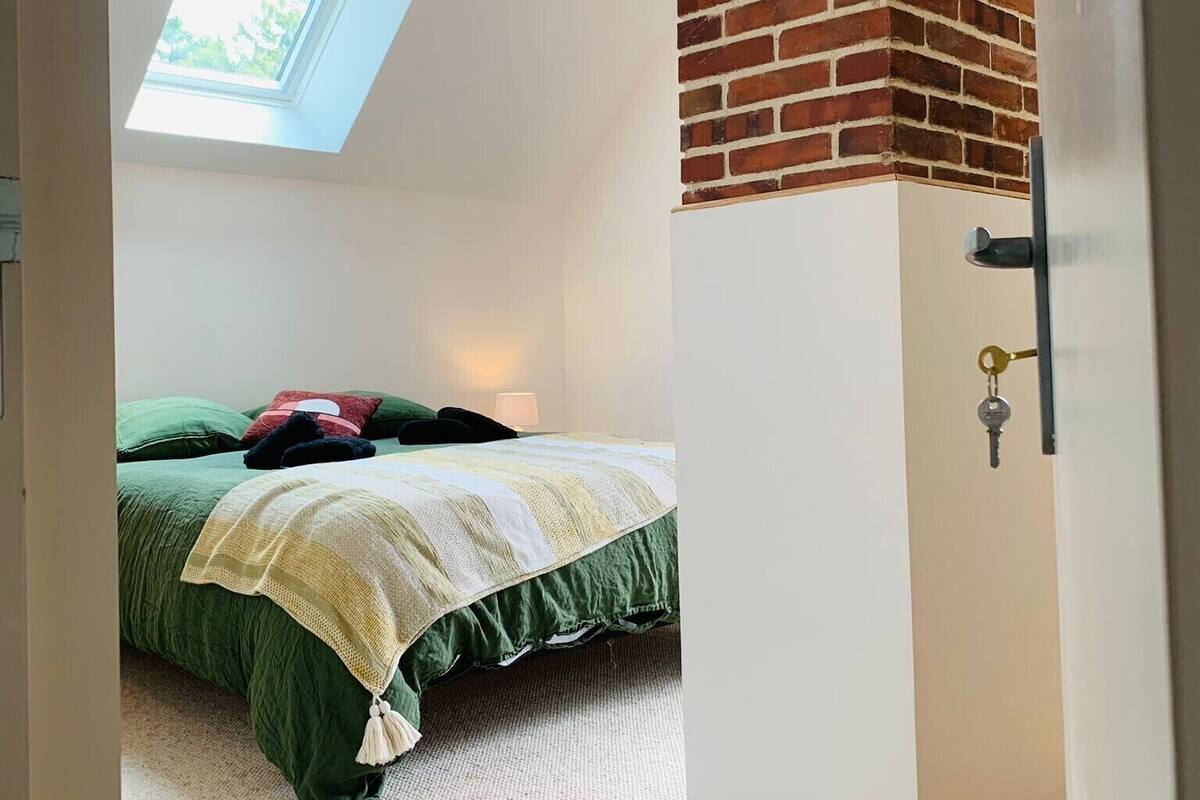 Quarto com claraboia, cama com roupa de cama verde, porta branca com chave e lareira de tijolos.
