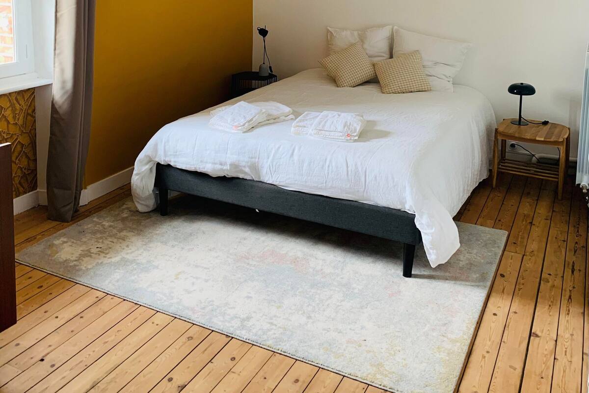 Quarto com cama, mesa de cabeceira, luminária, ventilador de teto e janela com cortinas.