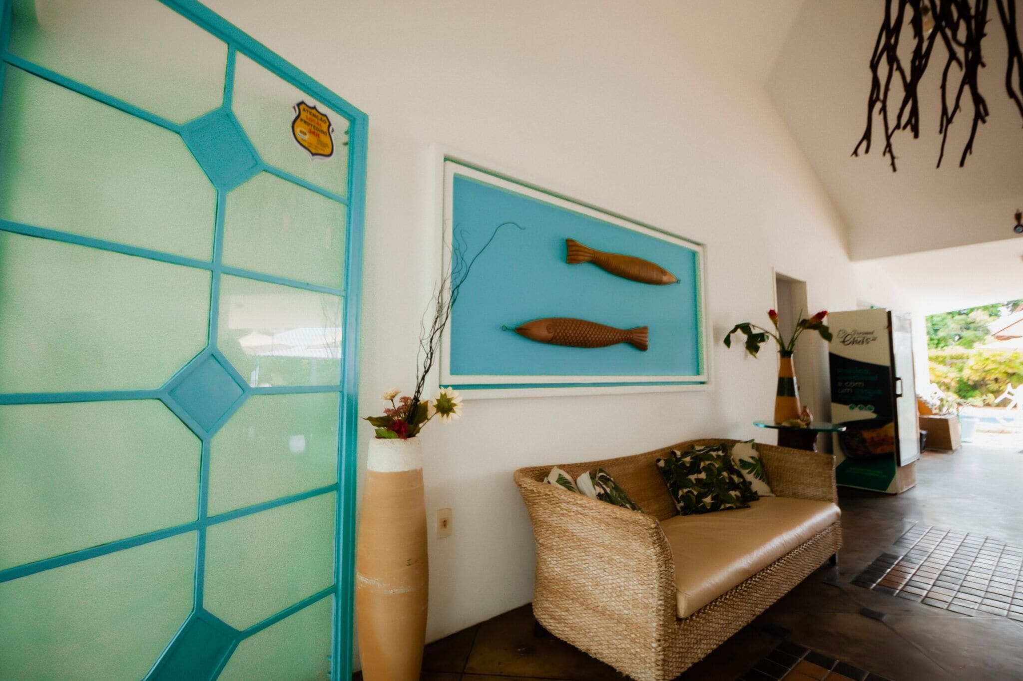 Quarto com uma janela ampla, um sofá de vime, um enfeite de peixe na parede e uma porta com padrão geométrico.