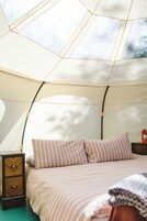 Quarto coberto por tenda, com uma cama, almofadas listradas, uma pequena mesa de cabeceira de madeira e um candeeiro.