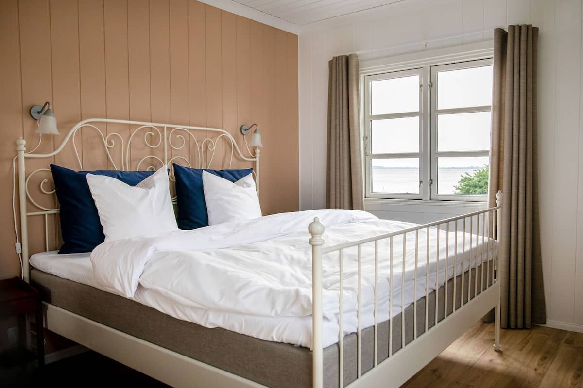Cama bem arrumada com roupa de cama branca e almofadas azuis, cabeceira de metal branca, e vista para o mar através de uma janela com cortinas.