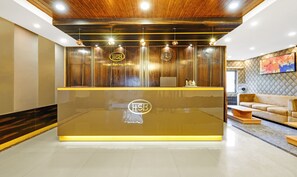 Bancada de recepção de hotel com acabamento em madeira e uma divisória de vidro. A área da recepção possui um relógio e uma pequena estátua na parede.
