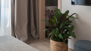 Quarto moderno com cama, televisão fixada na parede, planta em vaso e banheiro visível através de uma porta aberta.