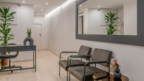 Uma área de espera moderna com balcão de recepção, duas cadeiras, um espelho grande e plantas em vasos.