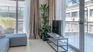 Uma sala de estar moderna com um sofá, uma televisão em um móvel, uma planta grande em vaso e uma porta de vidro deslizante que dá para uma varanda.