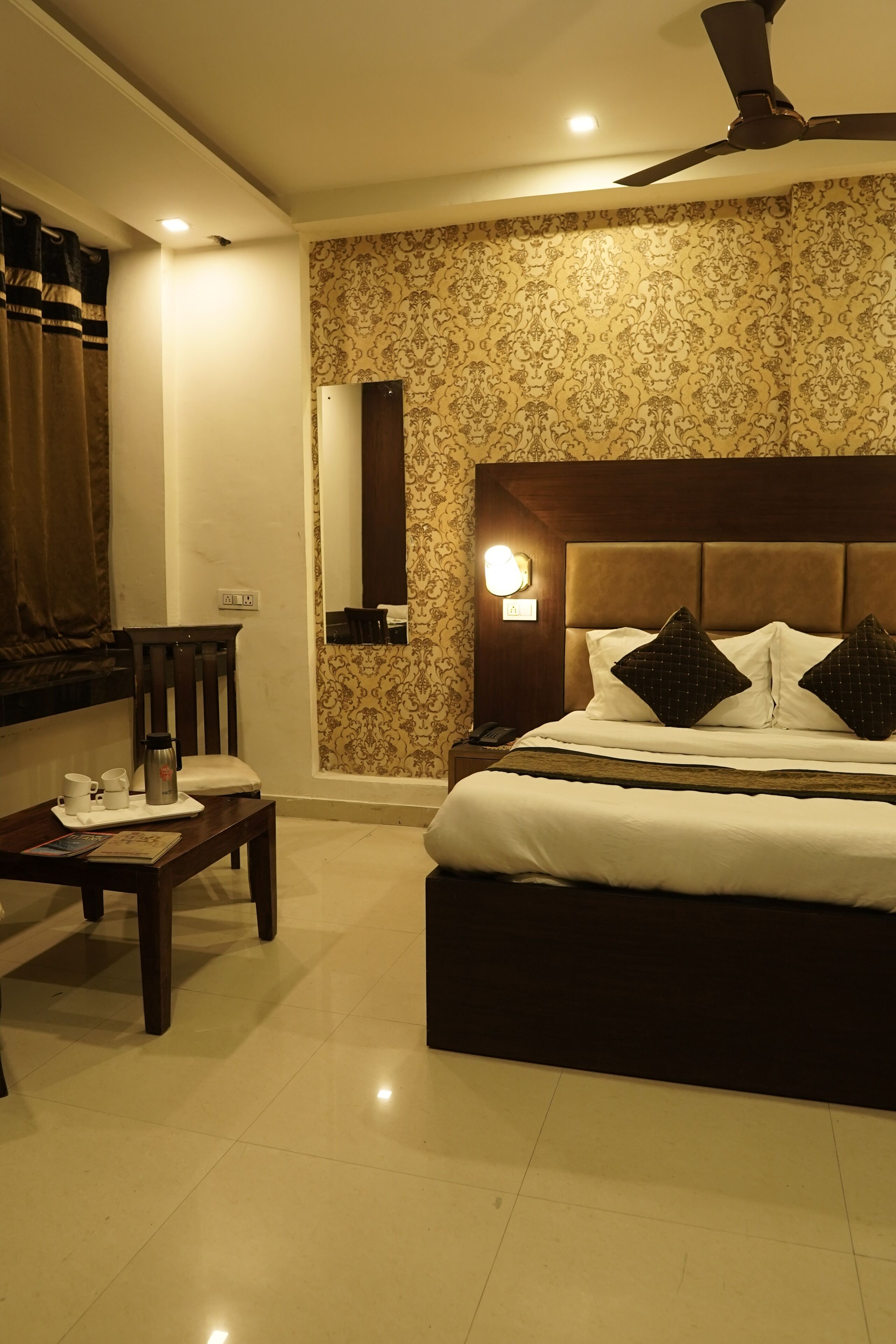 Quarto de hotel com uma cama grande, uma mesinha de cabeceira, uma cadeira, uma mesa com cafeteira e um ventilador de teto.