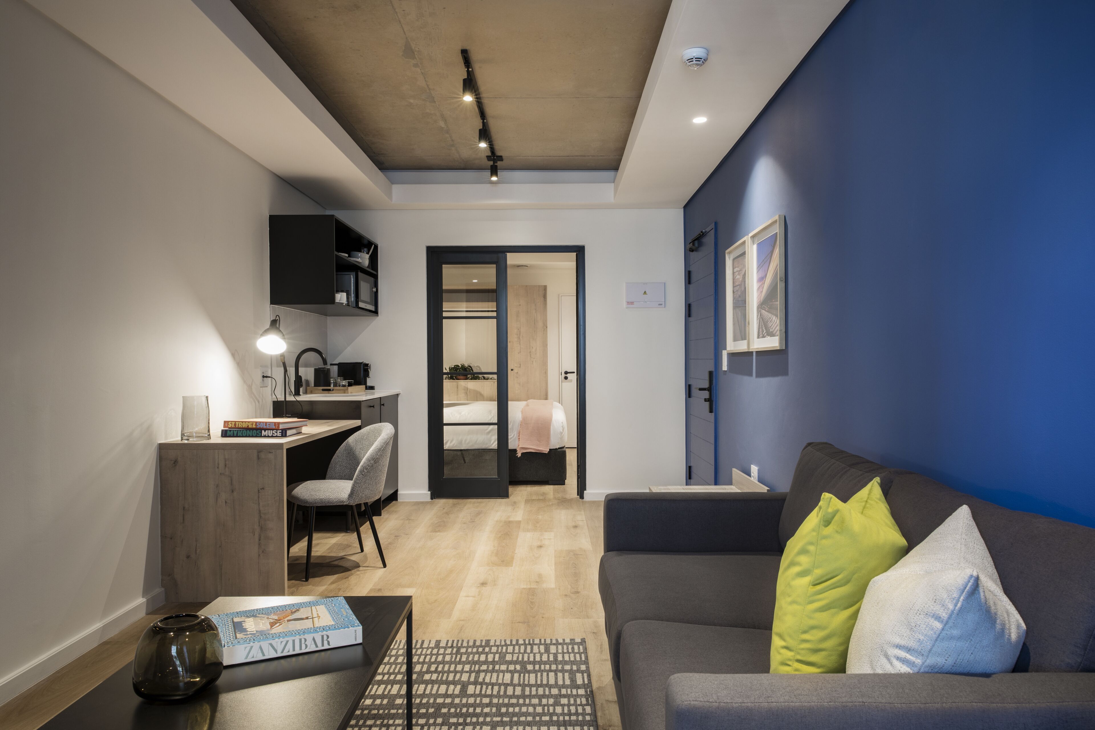 Uma sala de estar moderna com um sofá cinzento, uma mesa de centro e uma estante de livros. Há uma escrivaninha com uma cadeira e um abajur. O quarto tem um chão de madeira e uma parede azul de destaque.