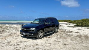 Um SUV preto estacionado numa praia de areia, com um céu azul límpido e oceano ao fundo.