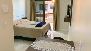 Quarto de hotel com cama, mesa com toalha de renda, luminária pendente e vista para a varanda externa.