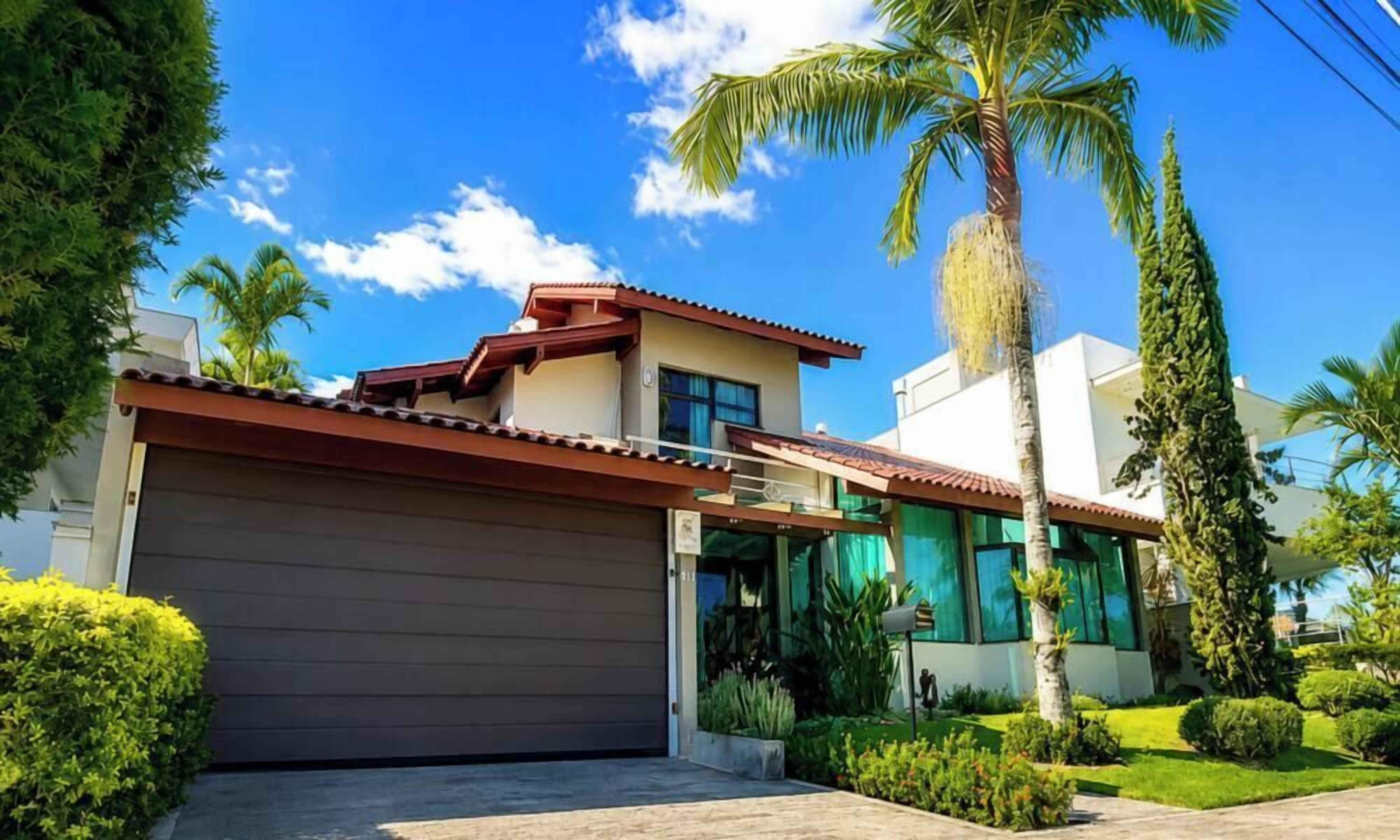 Uma casa moderna de dois andares com garagem, cercada por vegetação bem cuidada e palmeiras.