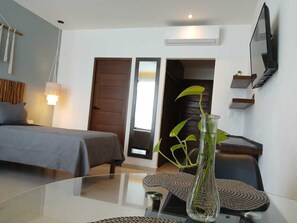 Quarto de hotel moderno com cama, uma mesinha com um vaso, uma cadeira, TV fixada na parede e uma porta que dá para outro quarto.