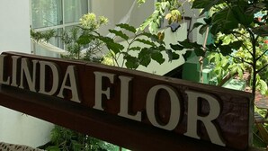 Um letreiro com a inscrição 'LINDA FLOR' emoldurado em madeira, uma varanda com guarda-corpo de vidro e plantas verdes exuberantes.