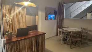 Um interior moderno com um balcão de recepção de madeira, um laptop, uma planta em vaso, uma mesa de jantar com cadeiras e uma escadaria ao fundo.